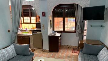 Um espaço compacto com sofá, uma mesinha com um rolo de papel higiênico, televisão na parede e área de cozinha com geladeira e pia.