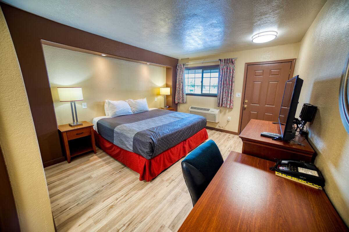 Quarto de hotel com cama, escrivaninha, cadeira, abajur e televisão.