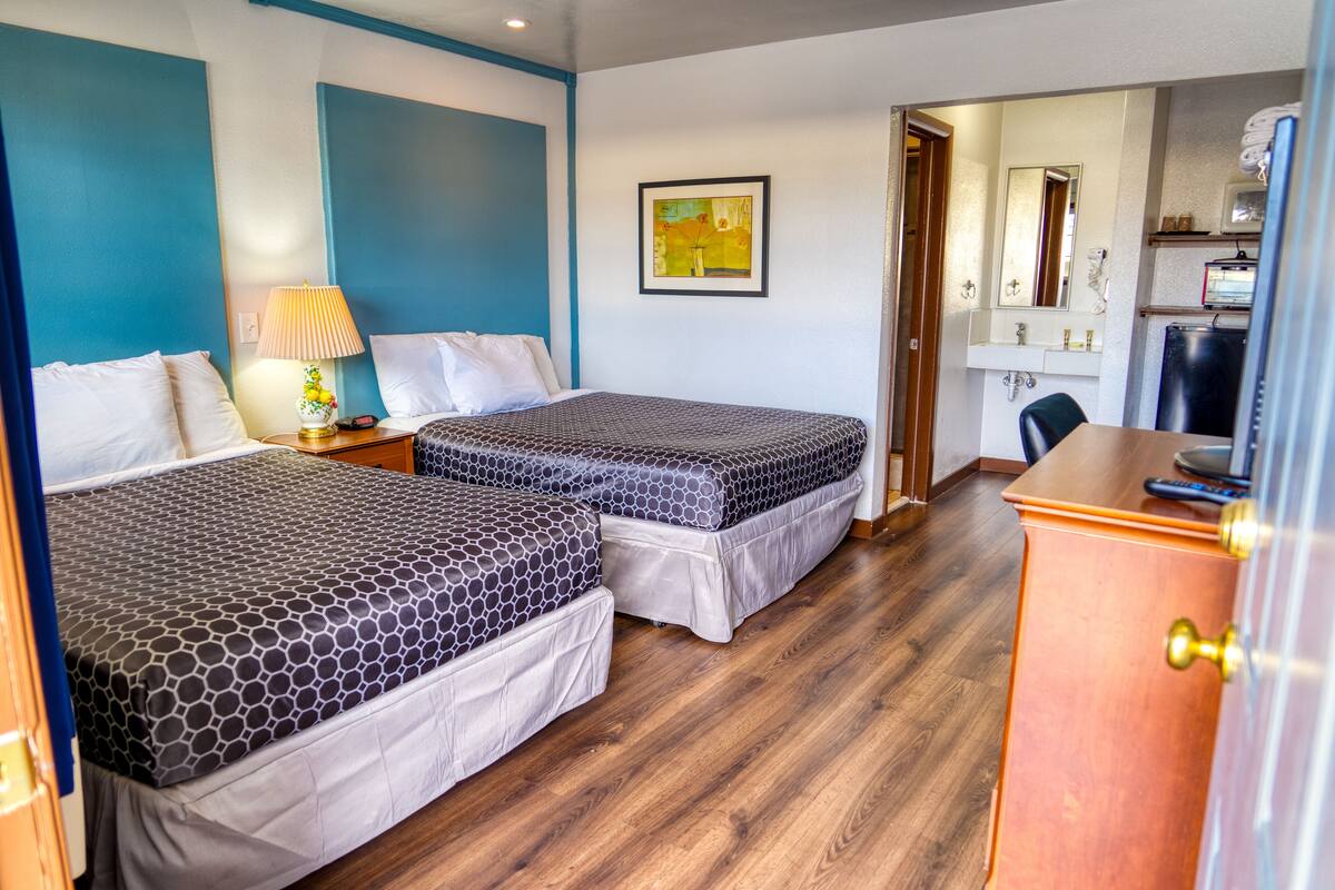 Quarto de hotel com duas camas, piso de madeira, uma mesa de trabalho e um banheiro visível por uma porta aberta.