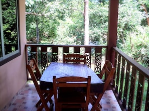 Uma mesa de madeira com toalha azul, rodeada por cadeiras, num varanda com vista para uma área arborizada.