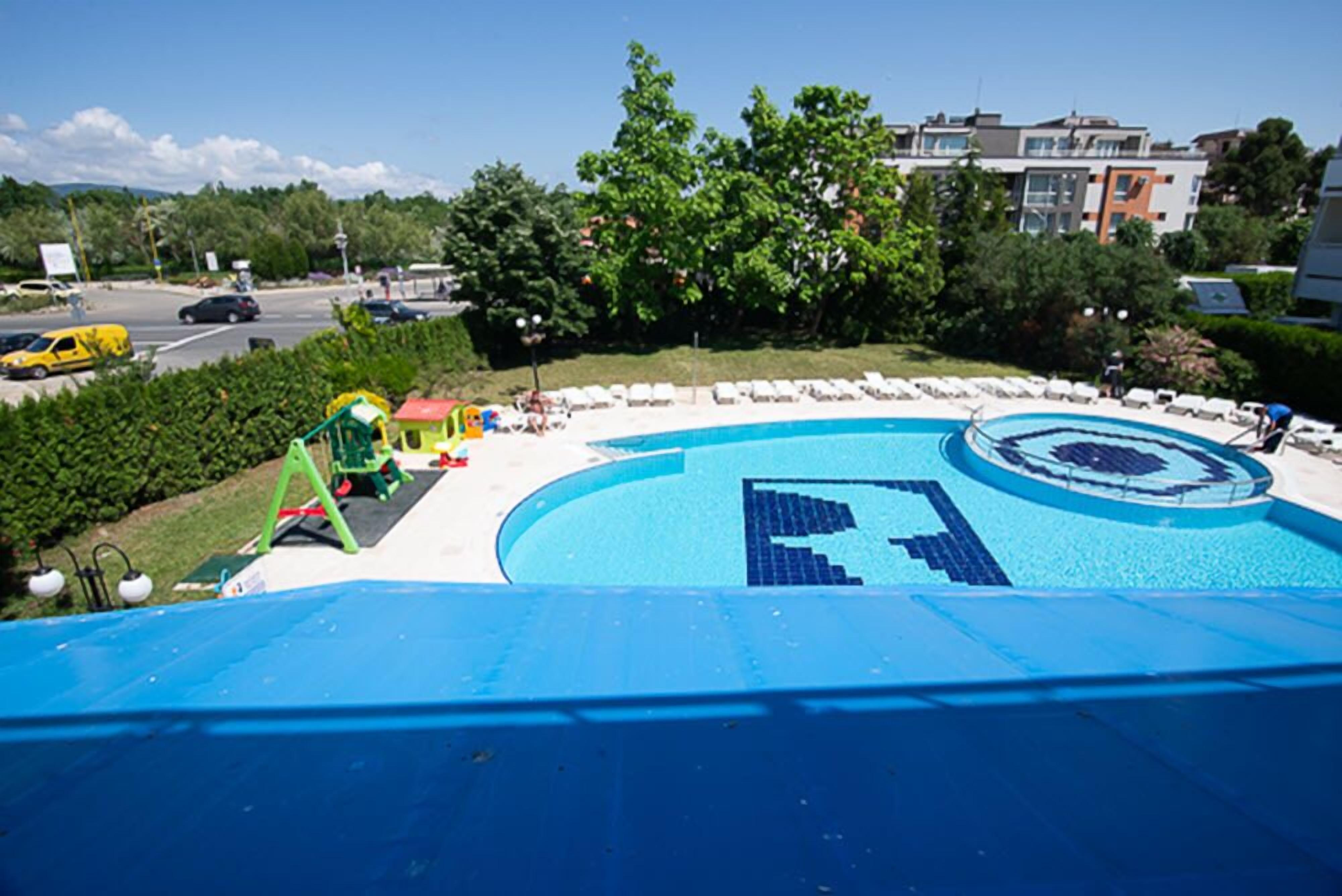 Piscina com deck azul, rodeada por vegetação e alguns edifícios ao fundo.