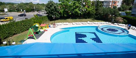 Piscina com deck azul, rodeada por vegetação e alguns edifícios ao fundo.