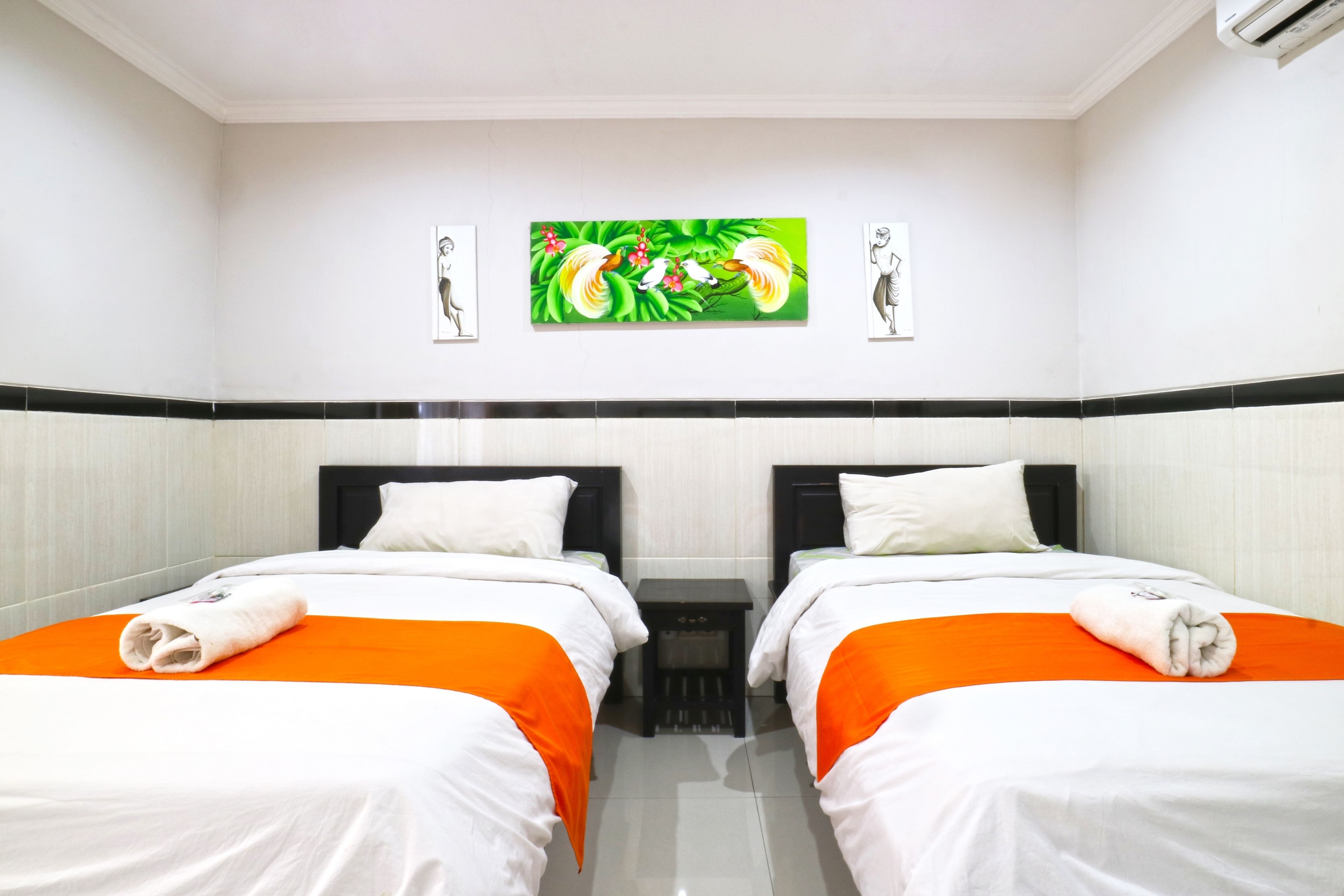 Quarto de hotel com duas camas, roupa de cama branca, colchas laranja, uma mesinha de cabeceira e um quadro pendurado na parede.