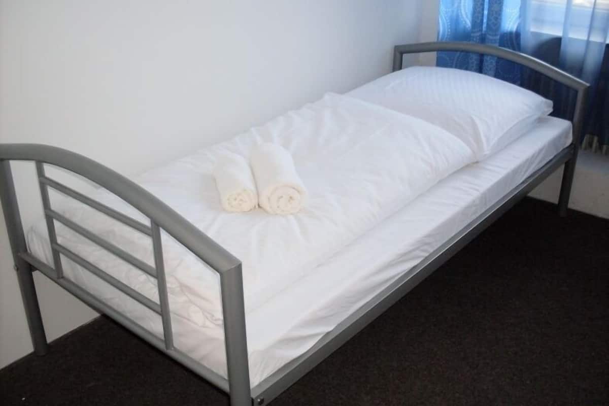 Uma cama de solteiro com estrutura metálica, roupa de cama branca e duas toalhas enroladas.