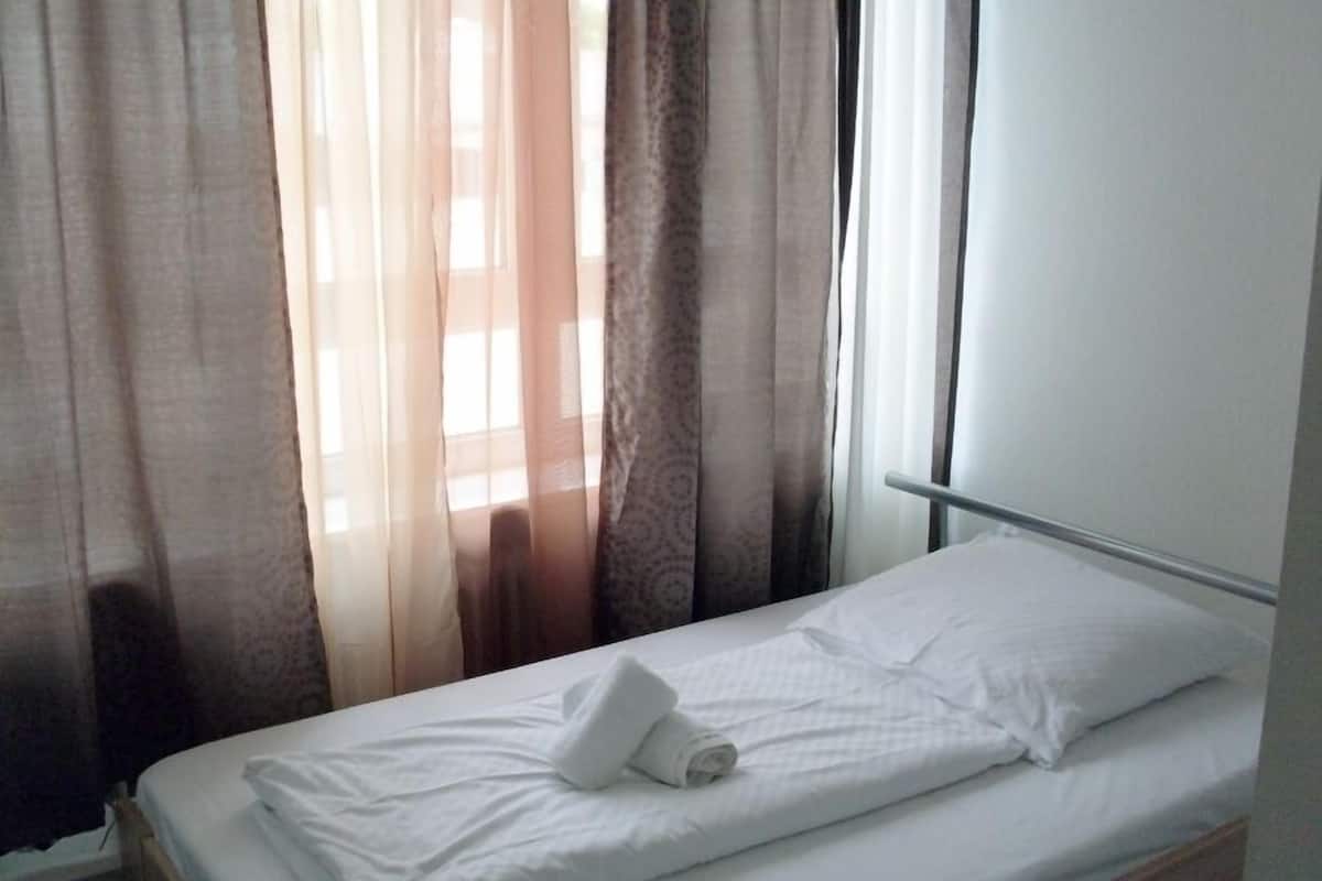 Uma cama de solteiro com roupa de cama branca, uma toalha enrolada, estrutura de metal e cortinas leves que permitem a entrada de luz natural.