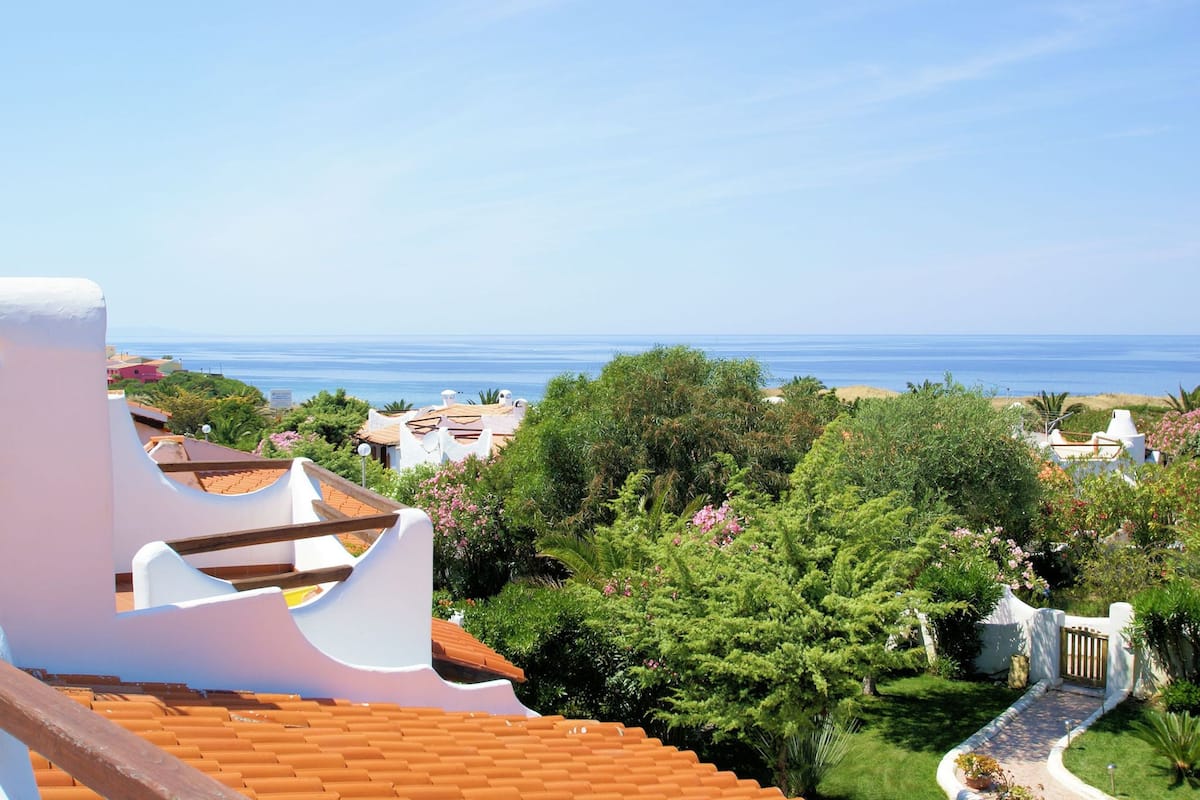 Vista costeira com telhados em tons de terracota, vegetação exuberante e céu azul límpido.