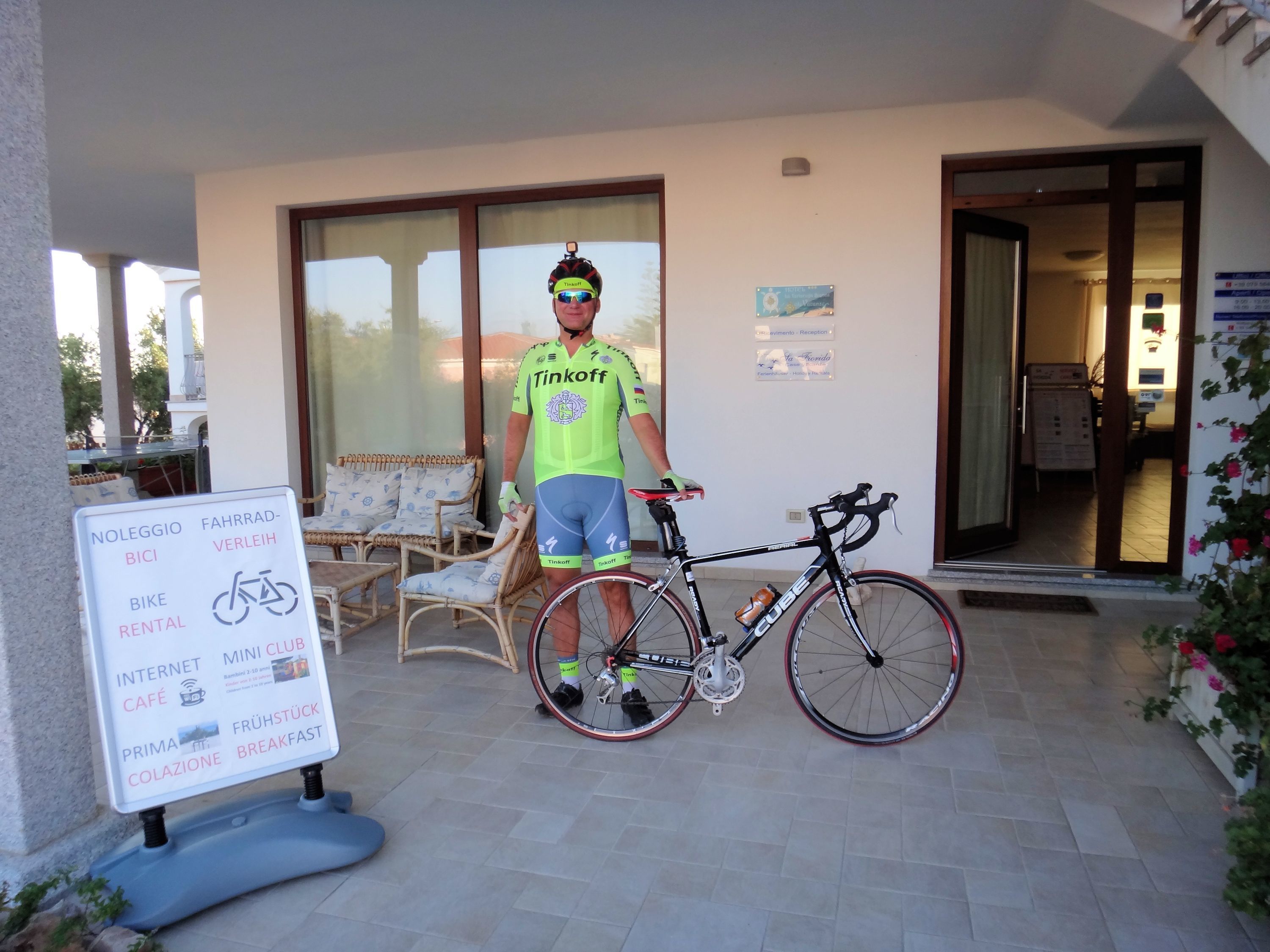 Uma pessoa vestida para andar de bicicleta está ao lado de uma bicicleta, com uma placa que lista comodidades como aluguel de bicicletas e café da manhã.