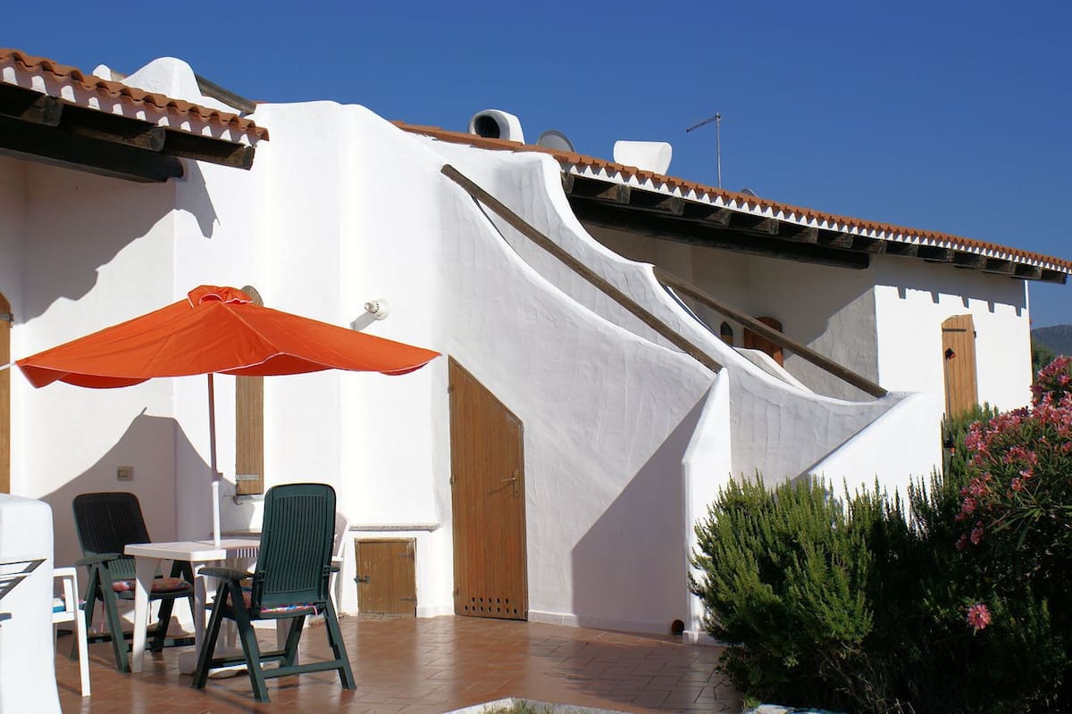 Uma área de estar externa com mesa e cadeiras, um guarda-sol vermelho e um edifício branco com telhado inclinado.
