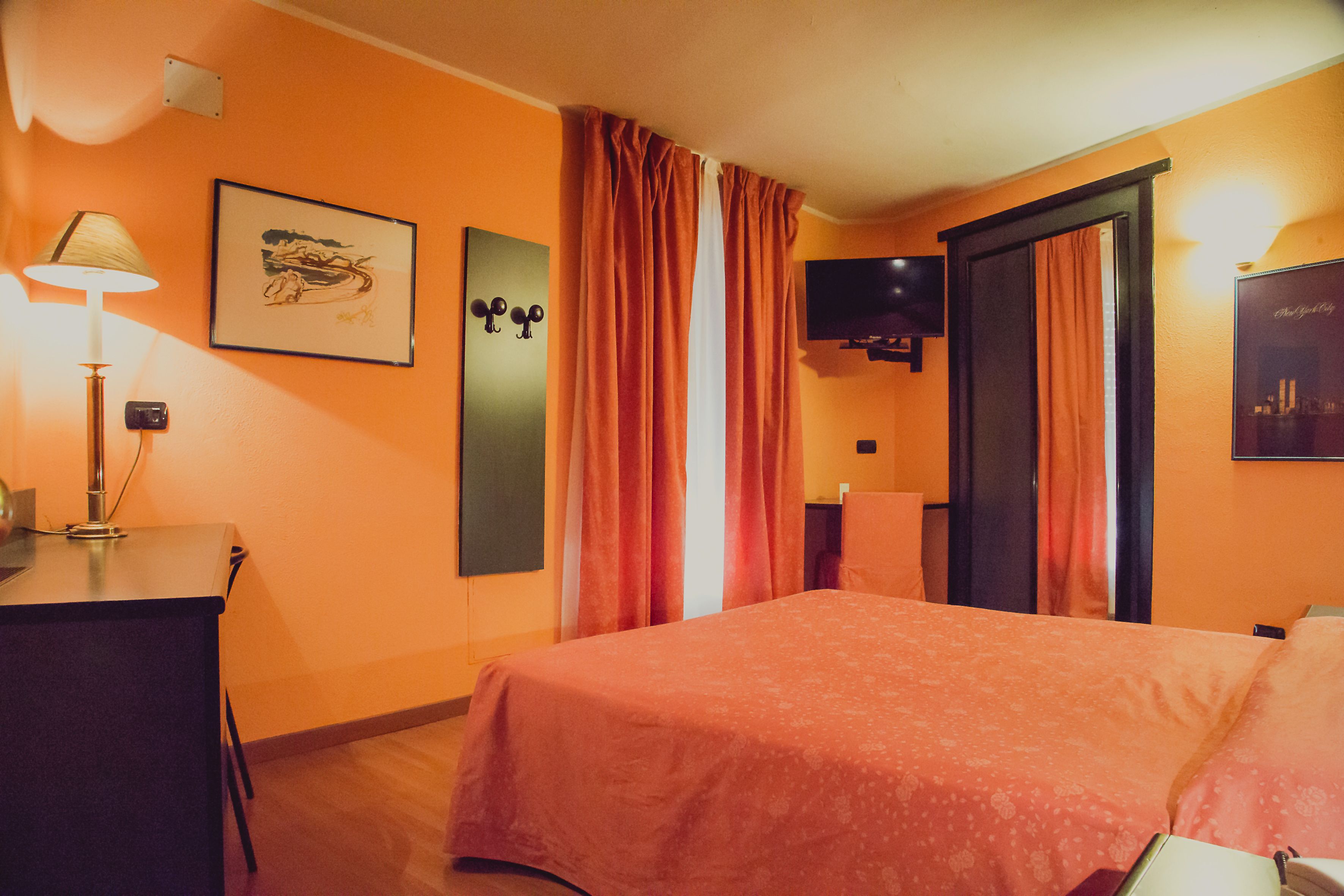 Quarto de hotel com cama, escrivaninha, luminária, televisão e quadro na parede.