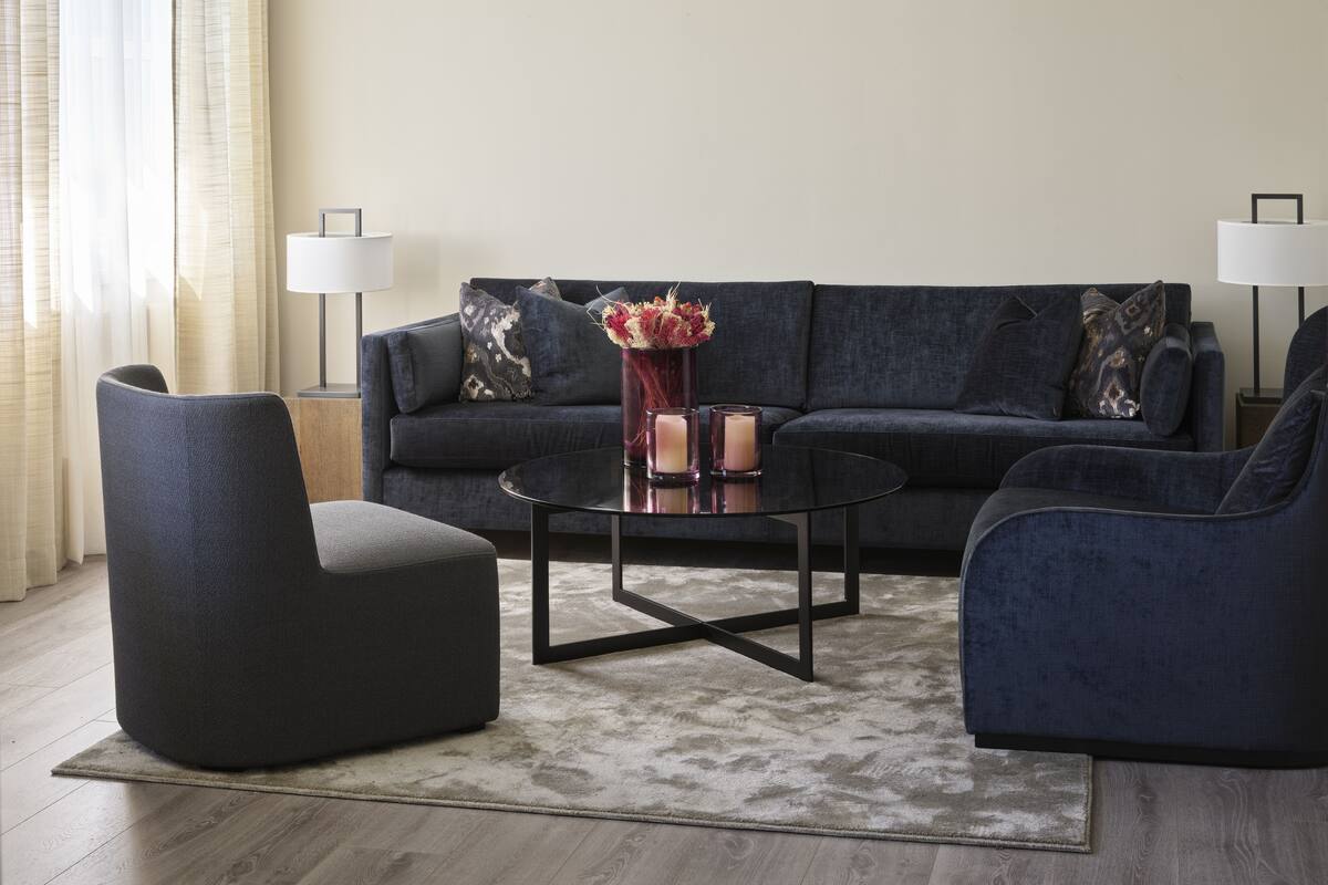 Uma sala de estar moderna com um sofá azul escuro, duas poltronas, uma mesa de centro de vidro e um vaso com flores.