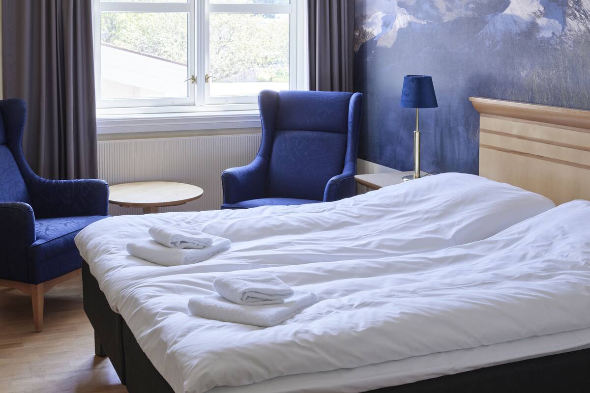 Quarto de hotel com uma cama grande, duas poltronas azuis, uma mesa redonda pequena e um abajur.