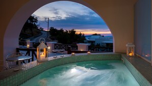 Uma banheira de hidromassagem com borda azulejada, cercada por uma varanda com cadeiras e mesas, e vista para o pôr do sol sobre uma estrada e árvores.