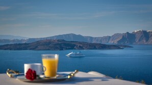 Uma mesa com um copo de suco de laranja, uma xícara branca e uma rosa vermelha. Ao fundo, um navio de cruzeiro visível na água, com montanhas à distância.