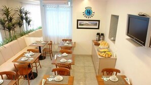 Uma sala de jantar de hotel com várias mesas preparadas para o pequeno-almoço, uma televisão e uma máquina de café fixada na parede.