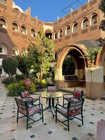 Um pátio com uma mesa de vidro redonda e quatro cadeiras, rodeado por um edifício de tijolos com arcos e um jardim.