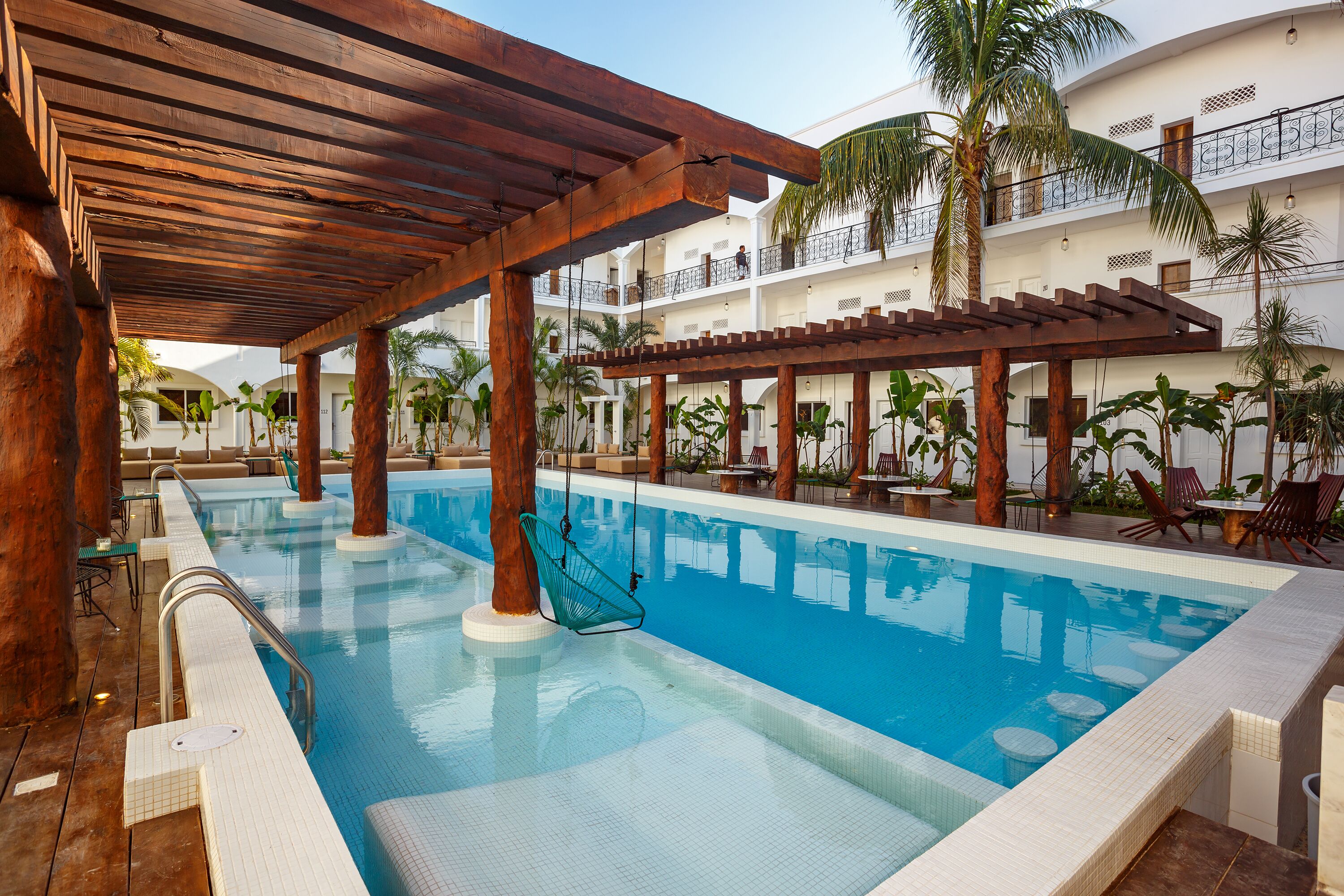 Área da piscina com pergolado de madeira, cadeiras de descanso e vista para um edifício com varandas.