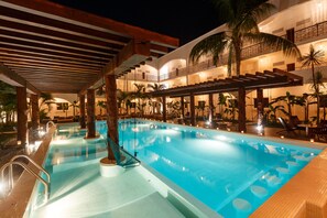 Uma grande piscina interna com um deque de madeira e espreguiçadeiras, rodeada por palmeiras e um edifício com vários andares e varandas.