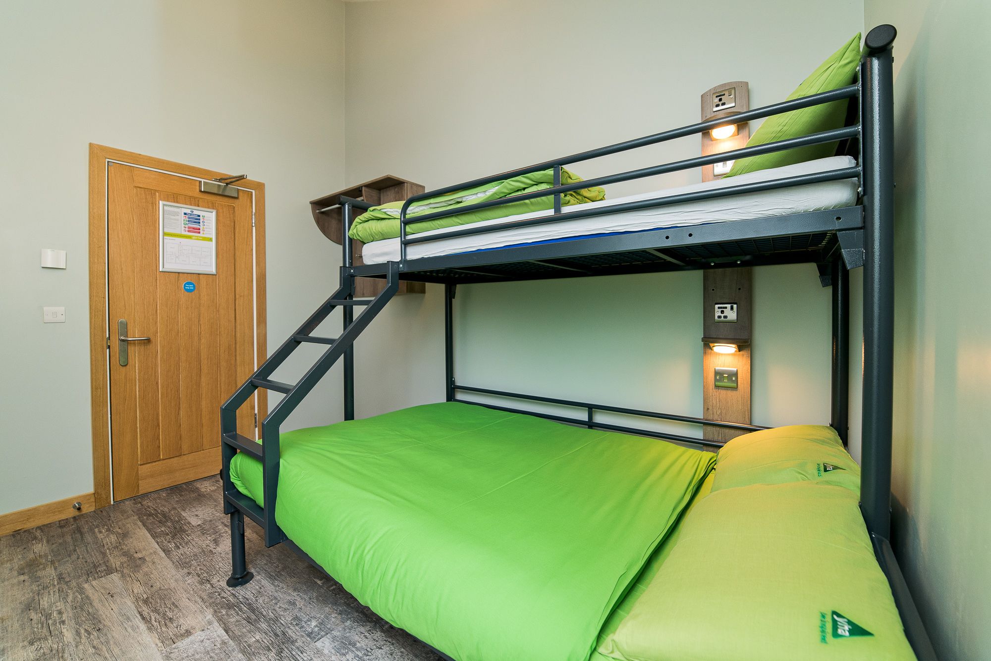 Uma cama beliche com roupa de cama verde, uma porta de madeira com um aviso e uma tomada na parede.