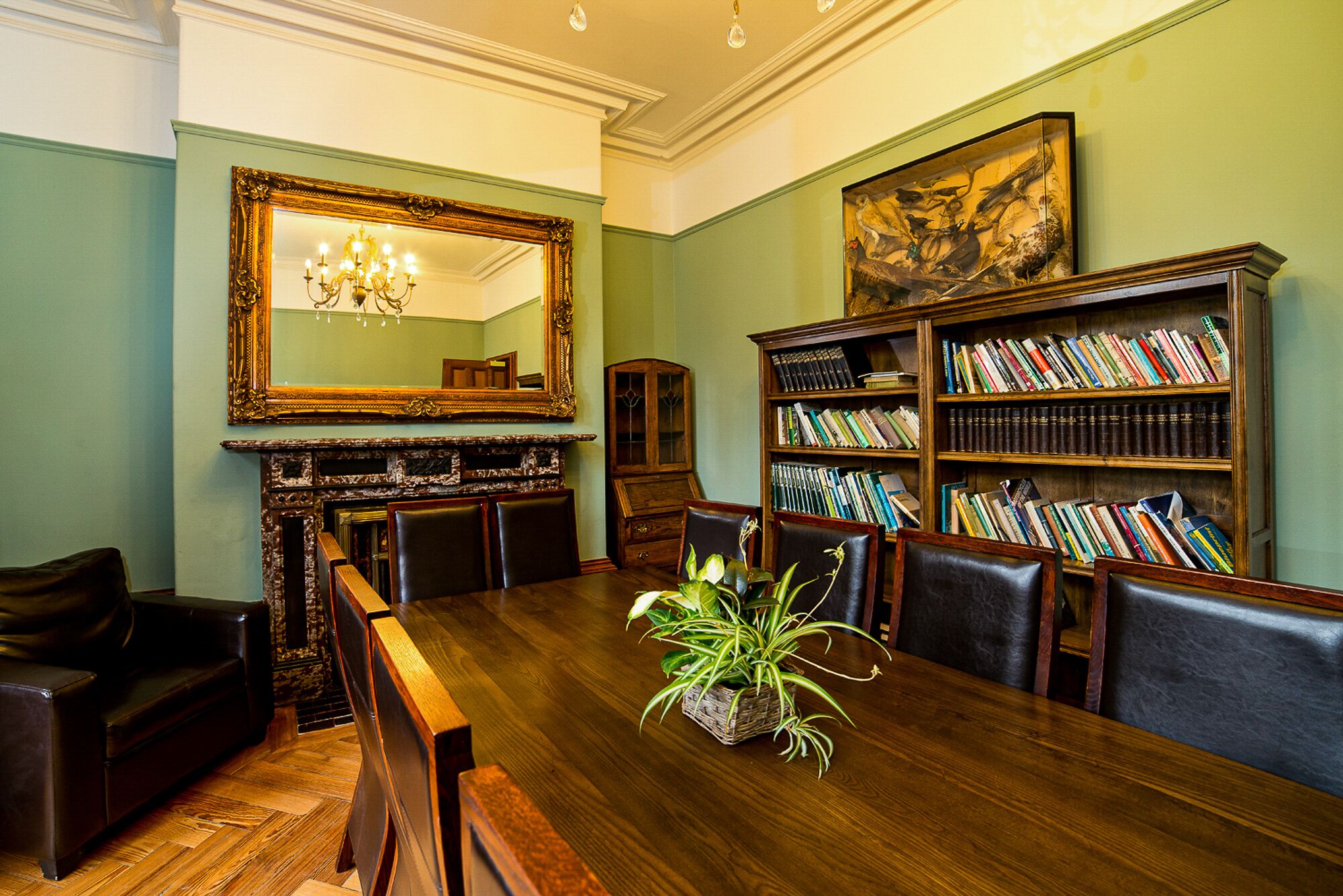 Uma sala de jantar formal com uma mesa de madeira, cadeiras, um candelabro, uma lareira, um espelho grande e uma estante de livros cheia.