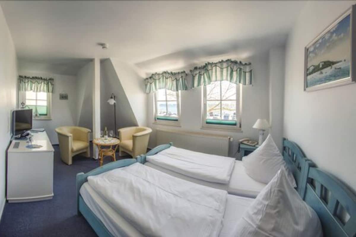 Quarto de hotel com duas camas de solteiro, uma escrivaninha com computador, duas cadeiras, uma mesinha e uma janela com cortinas.
