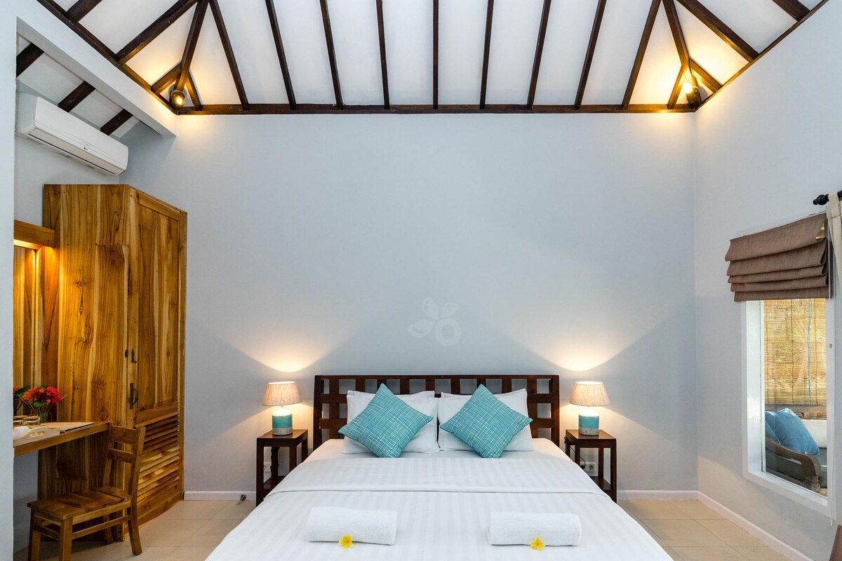 Quarto com cama, duas mesas de cabeceira com abajures, guarda-roupa de madeira, janela com cortinas e teto com vigas e luminárias de madeira.