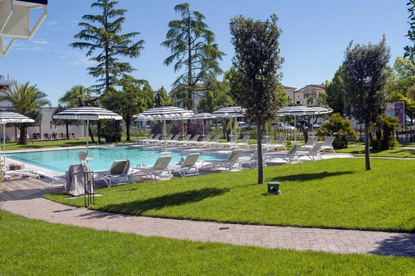 Uma área à beira da piscina com espreguiçadeiras e guarda-sóis, rodeada por gramado e árvores bem cuidados.