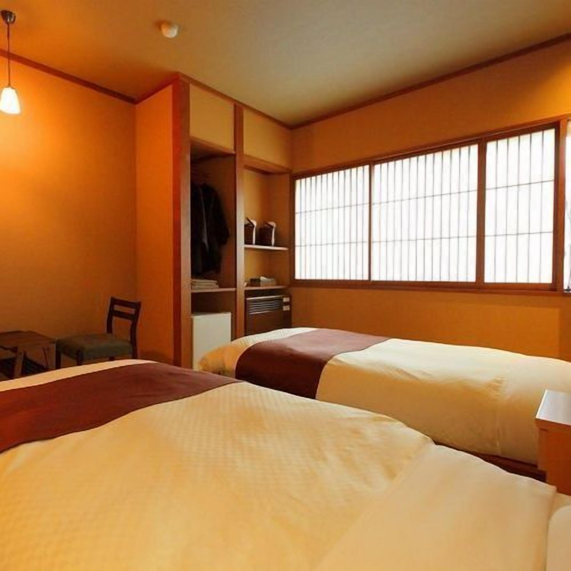 Quarto de hotel com duas camas, um guarda-roupa, uma secretária e uma janela com persianas verticais.