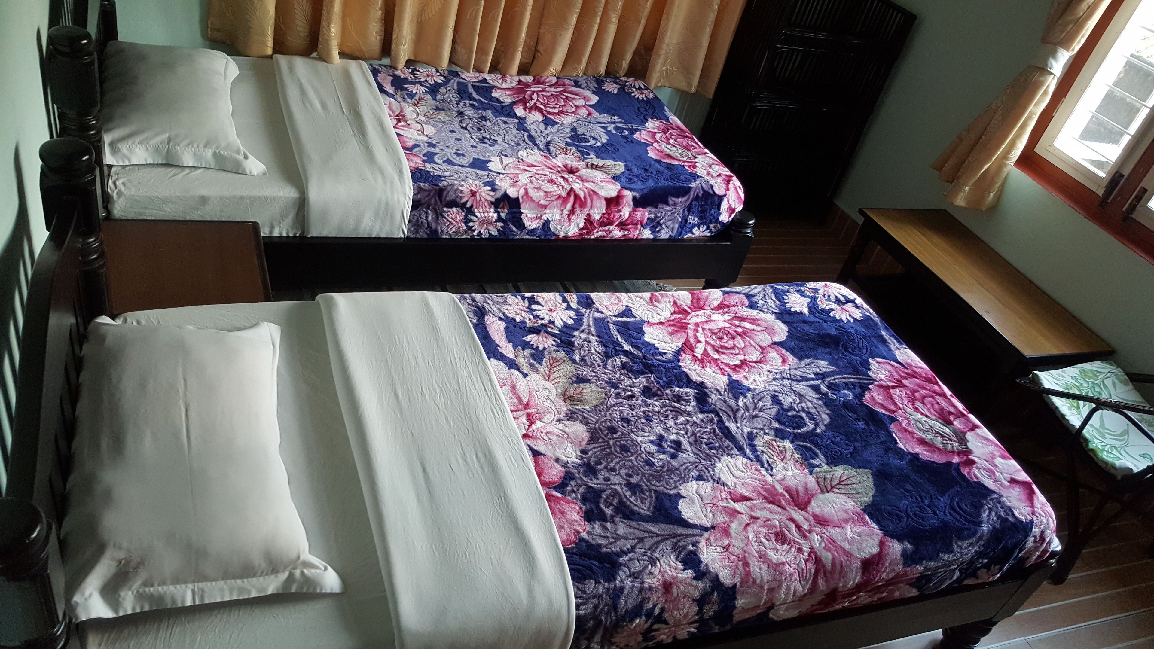 Quarto com duas camas, cada uma com colcha floral e almofadas brancas. Há um banquinho de madeira e uma mesinha.