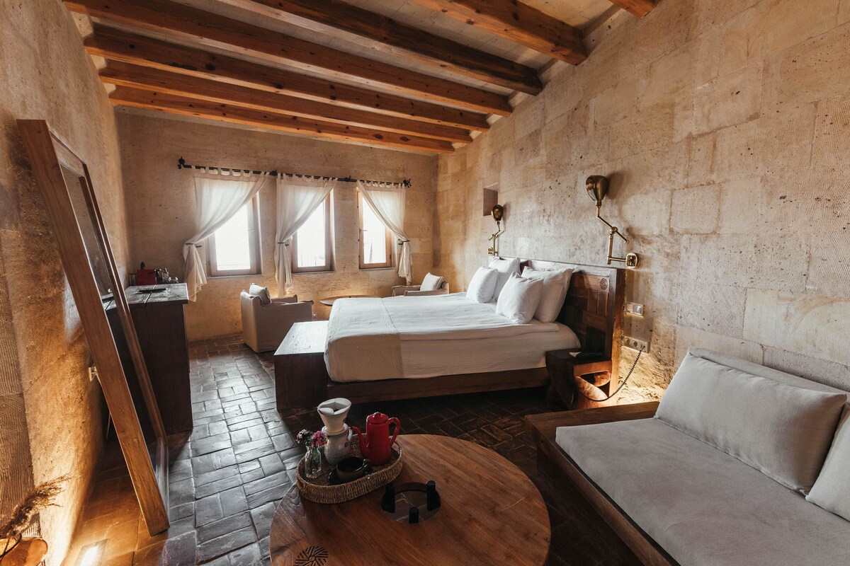 Um quarto acolhedor com teto de madeira, uma cama, um sofá, uma mesa pequena com bandeja e uma janela com cortinas.
