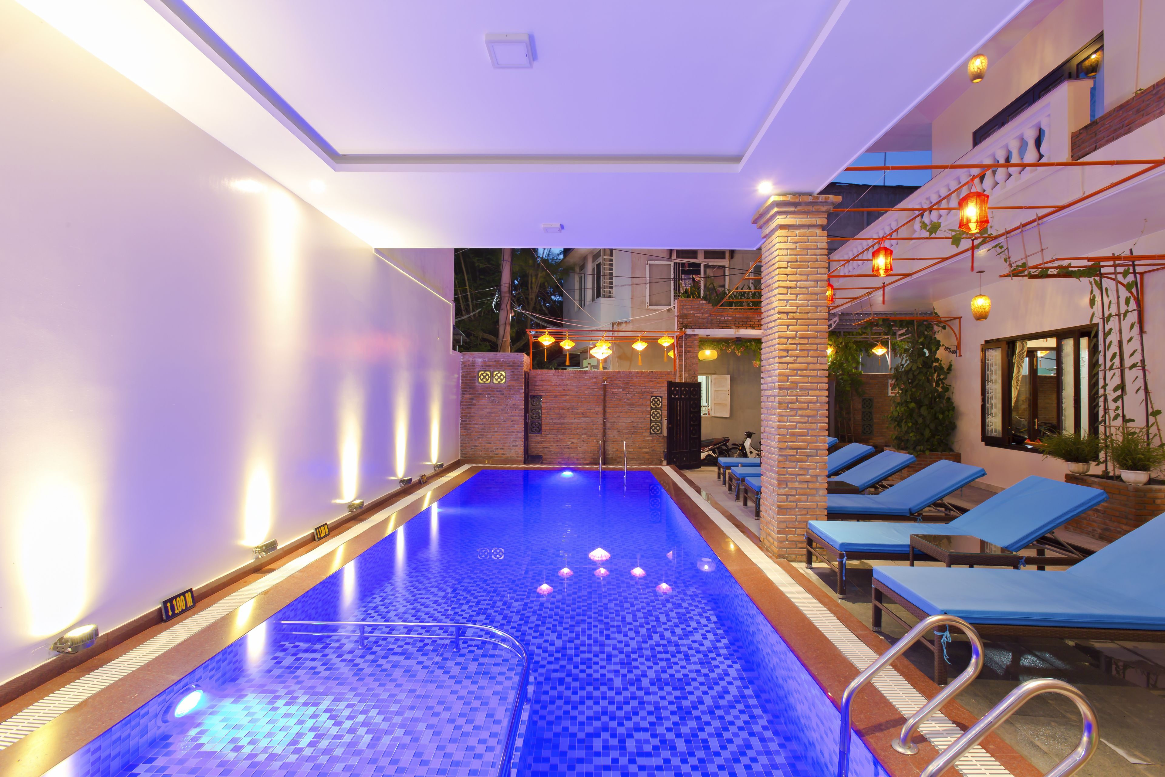 Piscina interna com piso de azulejos azuis, área de estar rebaixada e cadeiras de sol.