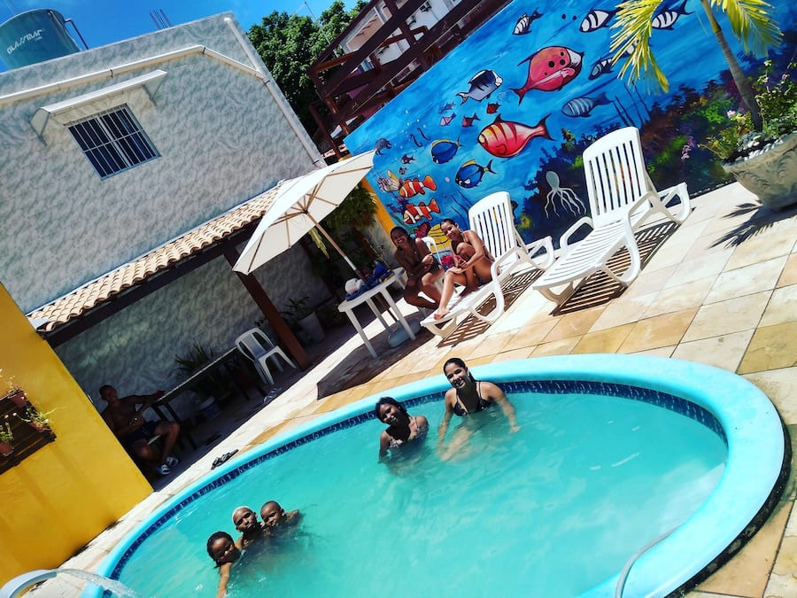 Piscina com um mural de peixes, rodeada por espreguiçadeiras brancas e um espaço de estar com piso cerâmico.