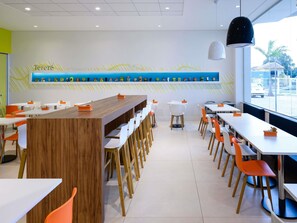 Um restaurante moderno com um balcão de madeira comprido, cadeiras brancas e laranja, e uma vitrine de comida na parede.