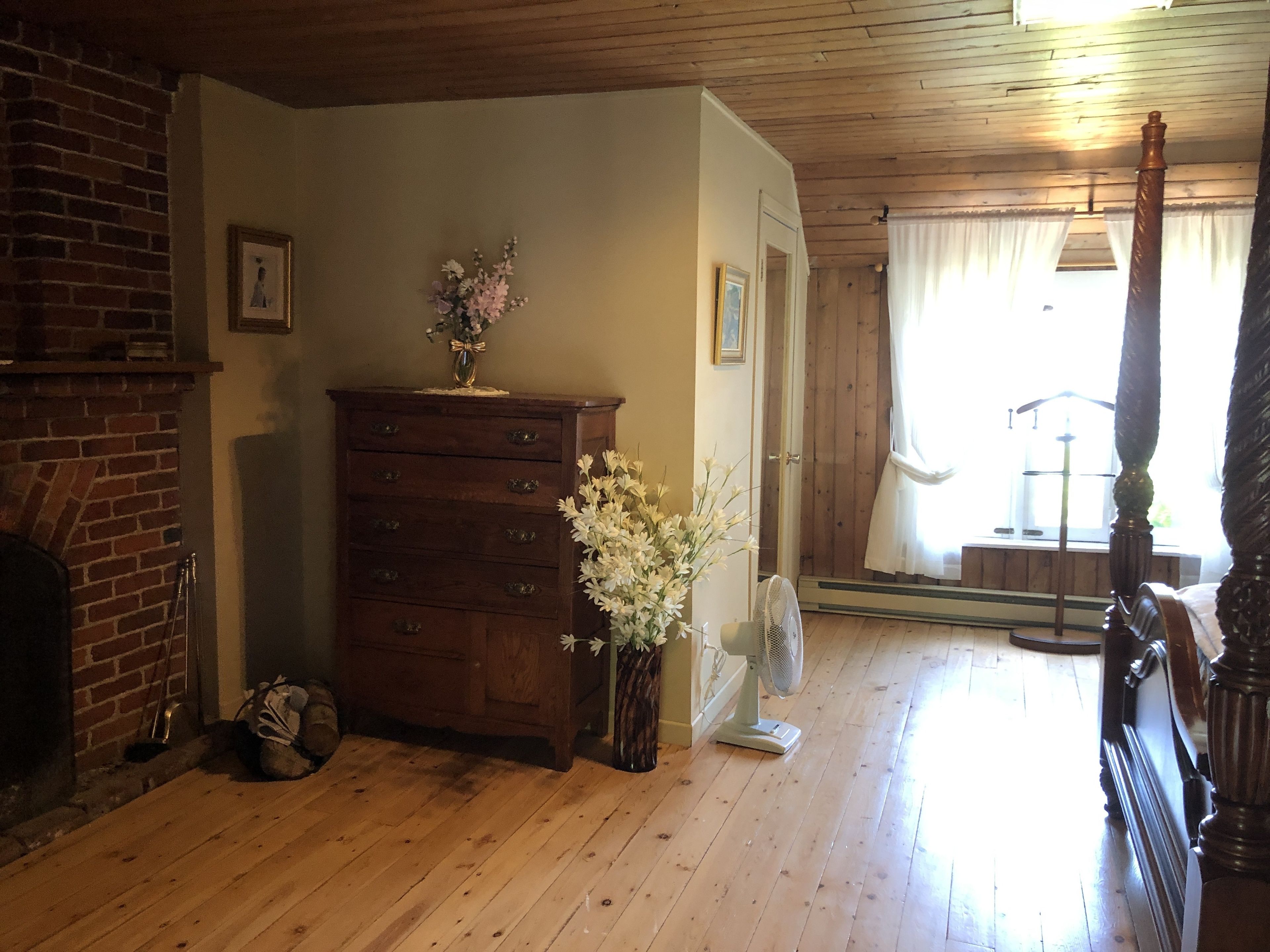 Quarto com piso de madeira, lareira, cômoda de madeira, ventilador e janela com cortinas.