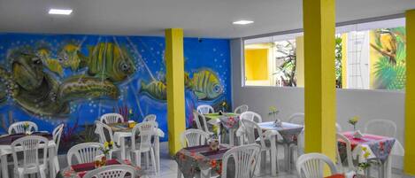 Restaurante com cadeiras e mesas de plástico branco, um mural de peixes na parede e pilares amarelos.