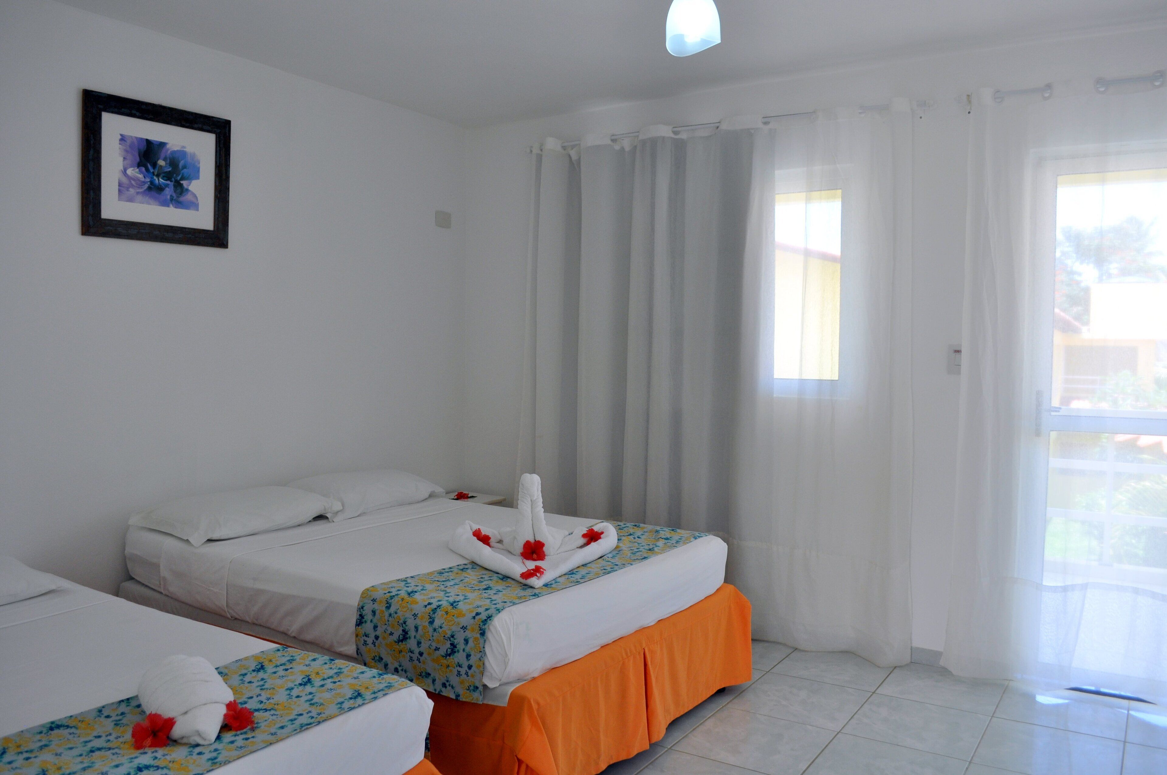 Quarto de hotel com duas camas, cada uma com um edredom branco e saia de cama laranja. Há um quadro na parede e uma janela com cortinas finas.