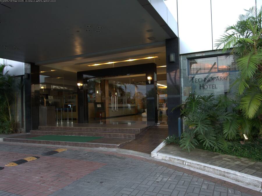 Entrada do Hotel Techron Park com fachada de vidro, carpete verde e área de jardim bem cuidada com palmeiras.