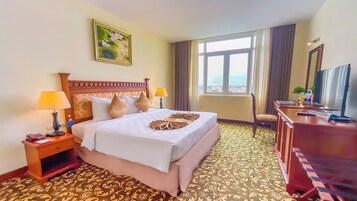 Quarto de hotel com uma cama grande, duas luminárias de cabeceira, uma escrivaninha com televisão e uma janela com cortinas.