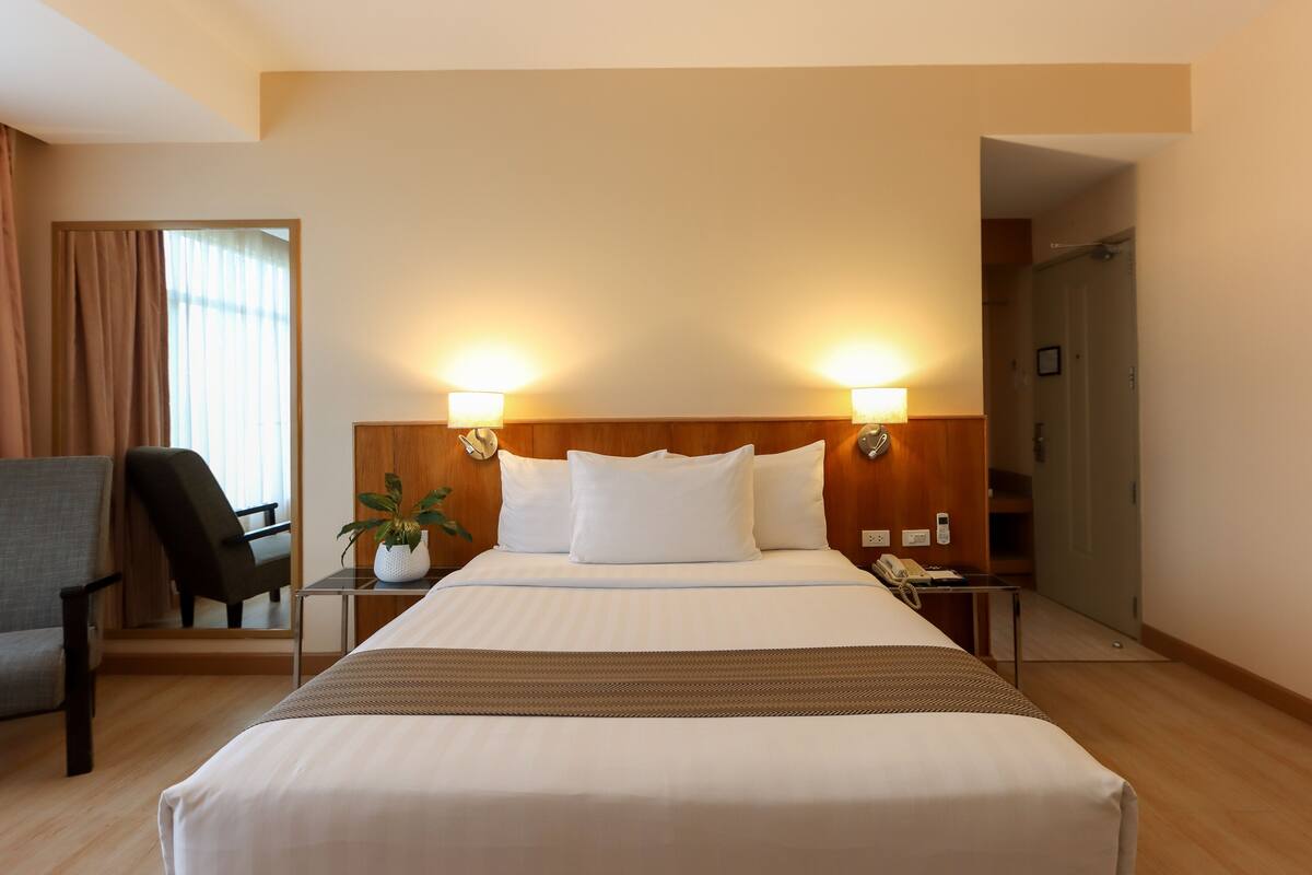 Quarto de hotel com uma cama grande, duas luminárias de cabeceira, uma cadeira, uma mesinha com uma planta e um telefone.