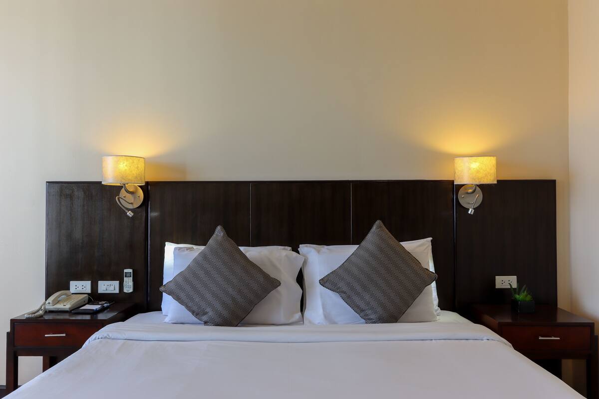Cama arrumada com duas almofadas, mesa de cabeceira com abajur e telefone no criado-mudo.