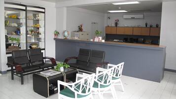 Uma área de recepção com balcão, duas cadeiras pretas, uma cadeira branca, uma mesinha com um globo e uma planta, e uma vitrine de vidro com flores e livros.