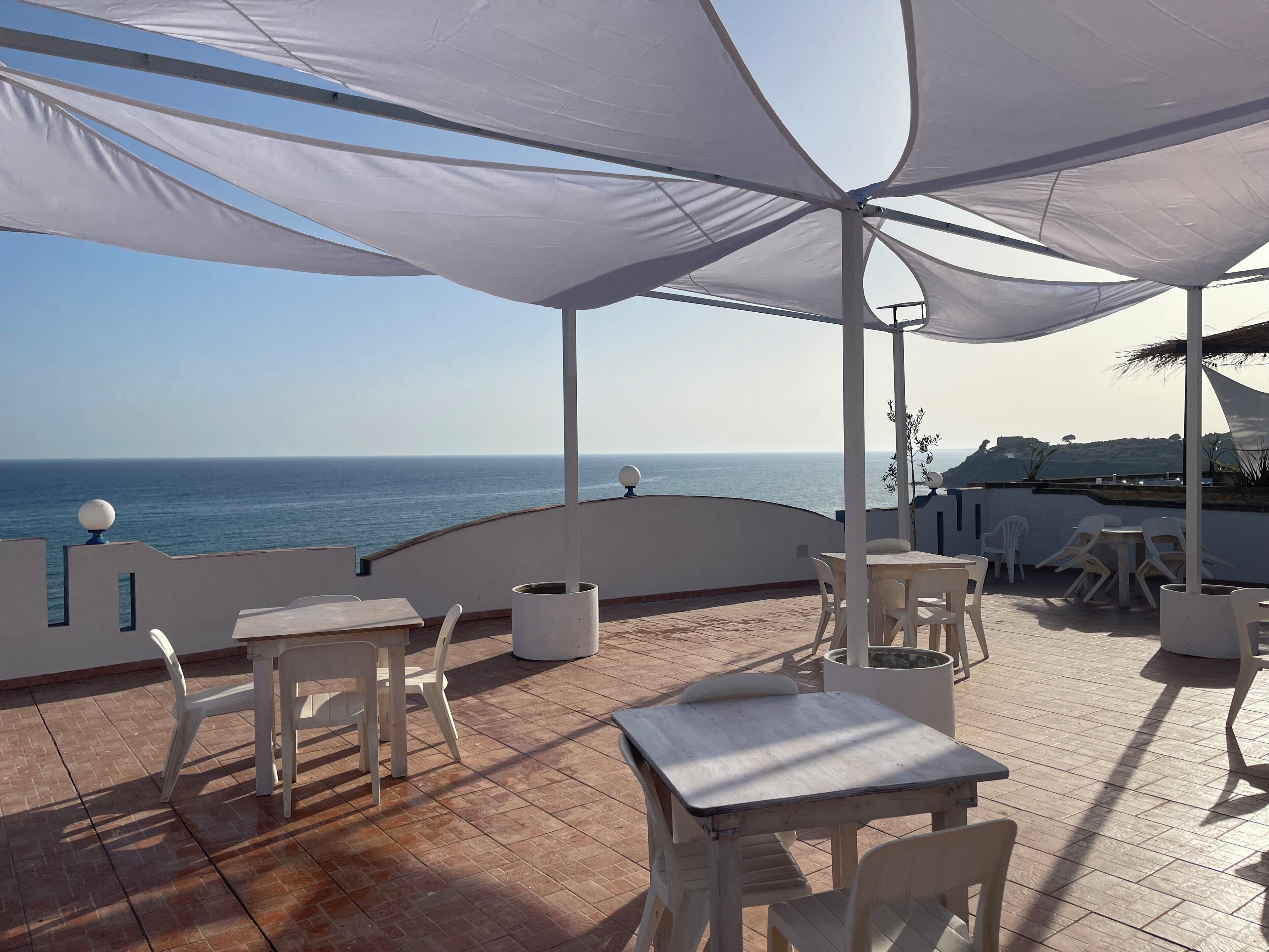 Terraço com mesas e cadeiras brancas, vista para o mar e uma estrutura de toldo branco.