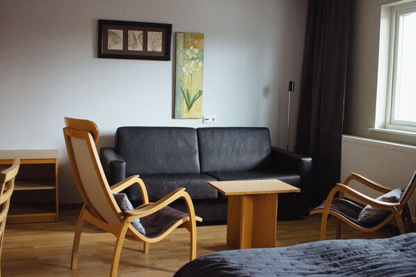Um quarto com um sofá de couro preto, duas cadeiras de madeira, uma mesa de madeira pequena e uma cama. Há quadros nas paredes e uma janela com cortinas.