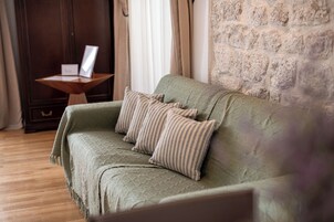 Um sofá verde com almofadas listradas em um quarto com piso de madeira, parede de pedra e uma pequena mesa de madeira com uma luminária.