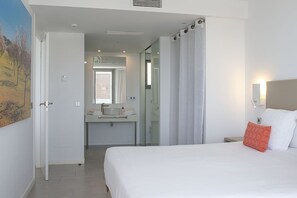 Quarto de hotel com cama, mesa de cabeceira, abajur, banheiro com pia e espelho, e porta que dá para outro quarto.