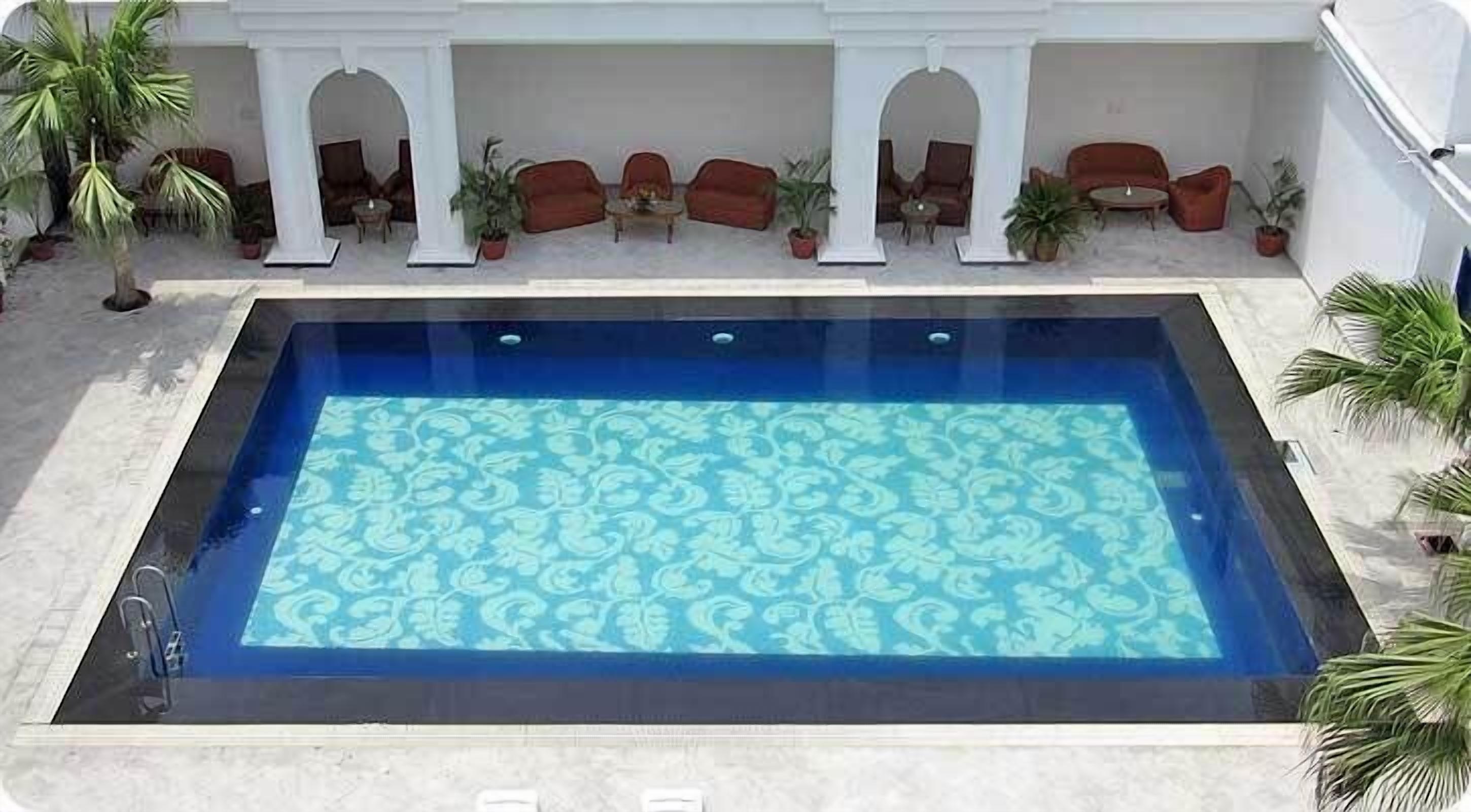 Uma piscina retangular com borda azul e um padrão floral na superfície da água, rodeada por uma estrutura branca com arcos e plantas em vasos.