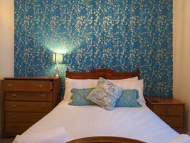 Uma cama bem arrumada com lençóis brancos e duas almofadas decorativas, uma mesa de cabeceira de madeira com abajur e uma cômoda com gavetas.