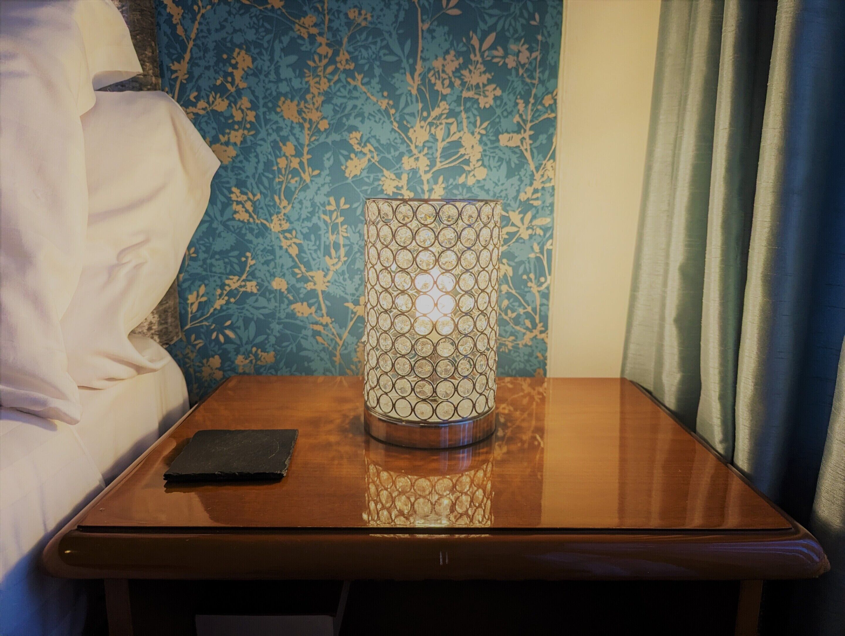 Uma mesa de cabeceira de madeira com uma luminária decorativa, um livro preto e uma cama com roupa de cama branca, tudo contra um papel de parede floral.