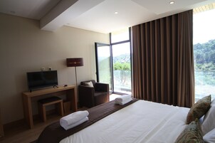 Quarto de hotel com uma cama grande, uma mesa com televisão, uma cadeira e vista para uma paisagem montanhosa através de uma janela ampla.