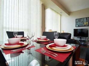 Uma área de refeições com mesa de vidro posta para dois, guardanapos vermelhos e um sofá de couro preto ao fundo.