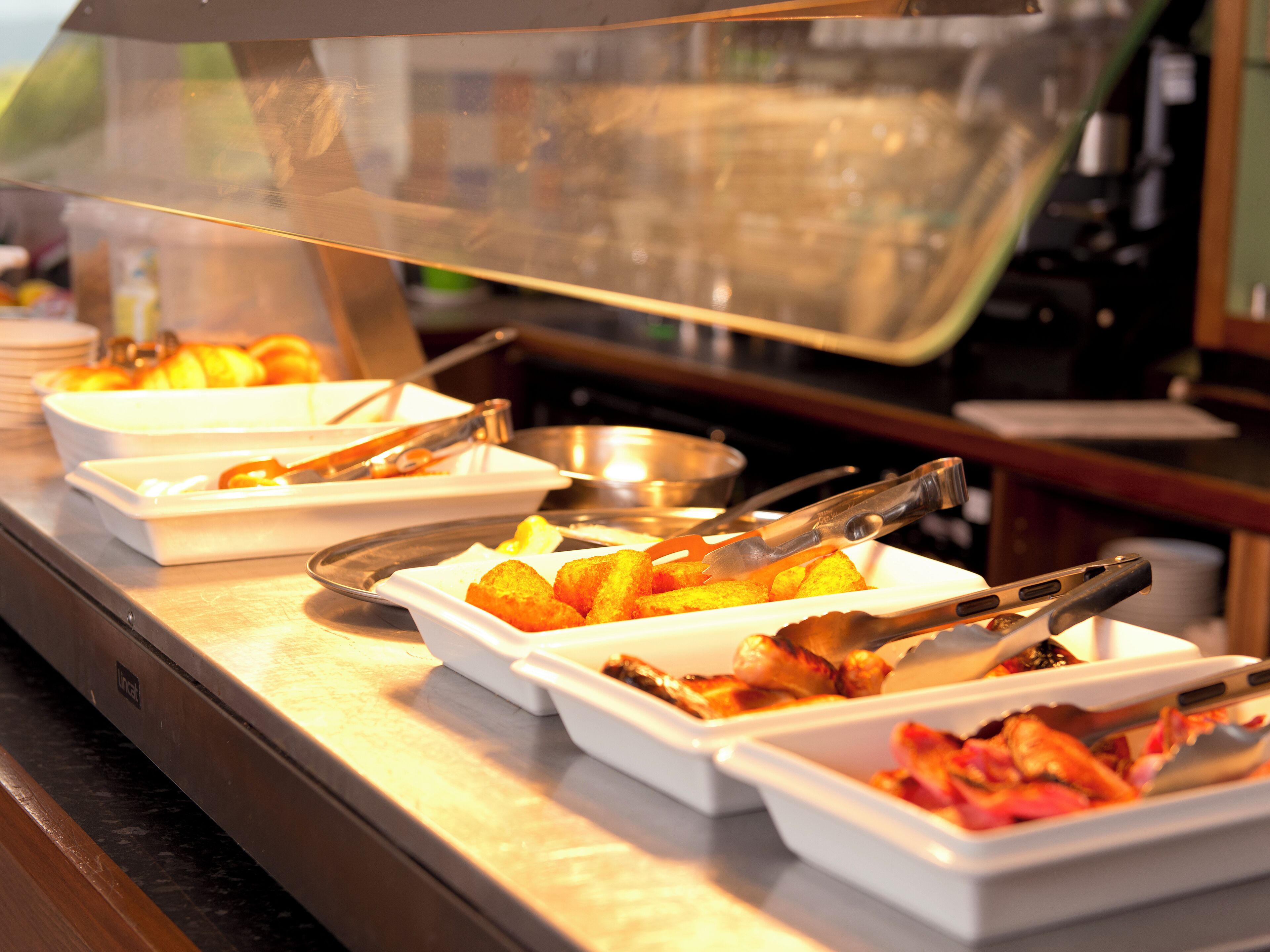Buffet com diversos pratos em bandejas brancas, incluindo frituras e carnes grelhadas.